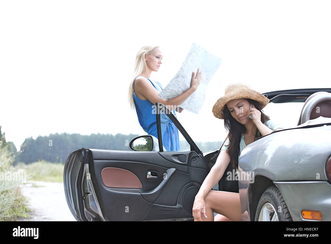 Woman using cell phone in convertible while friend reading map on road ...