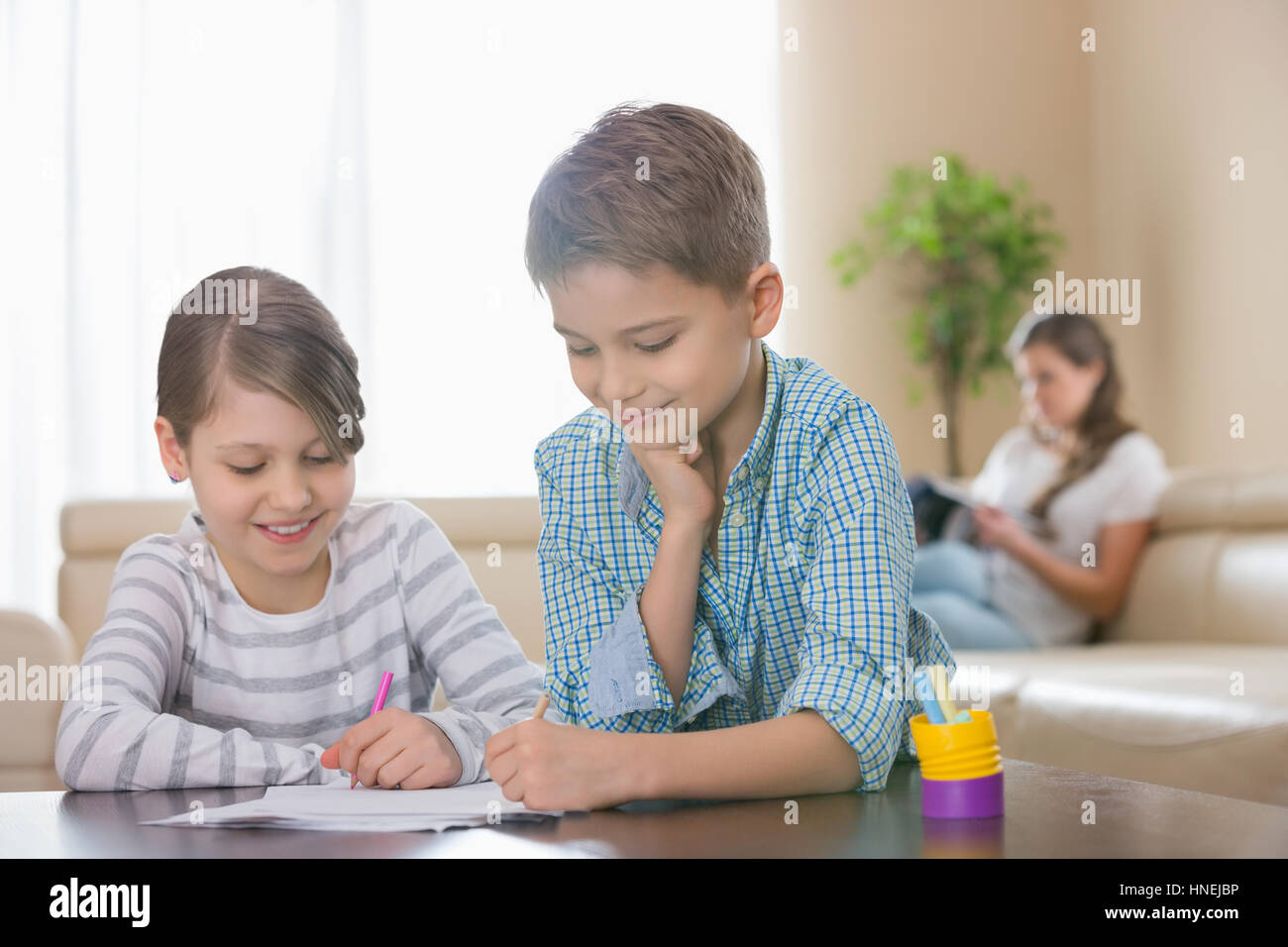 Siblings drawing together at table with mother in background Stock ...