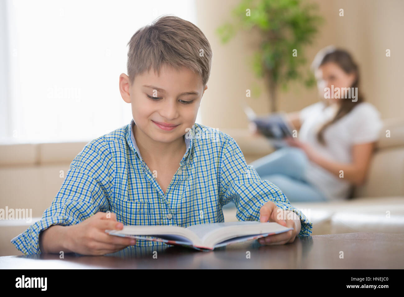 Cute boy reading book with mother in background at home Stock Photo - Alamy