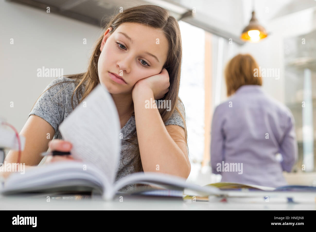 Bored girl studying at table with mother standing in background Stock ...