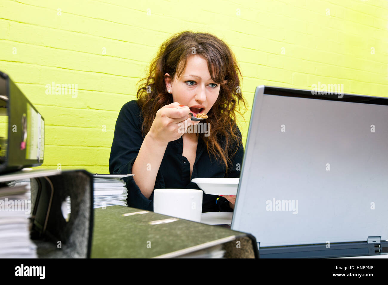 Caucasian young woman eating and working on her laptop computer at her ...