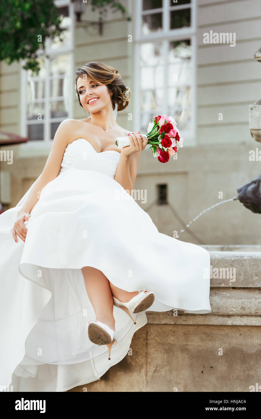 Nice bride sitting near monument and holding bouquet Stock Photo - Alamy