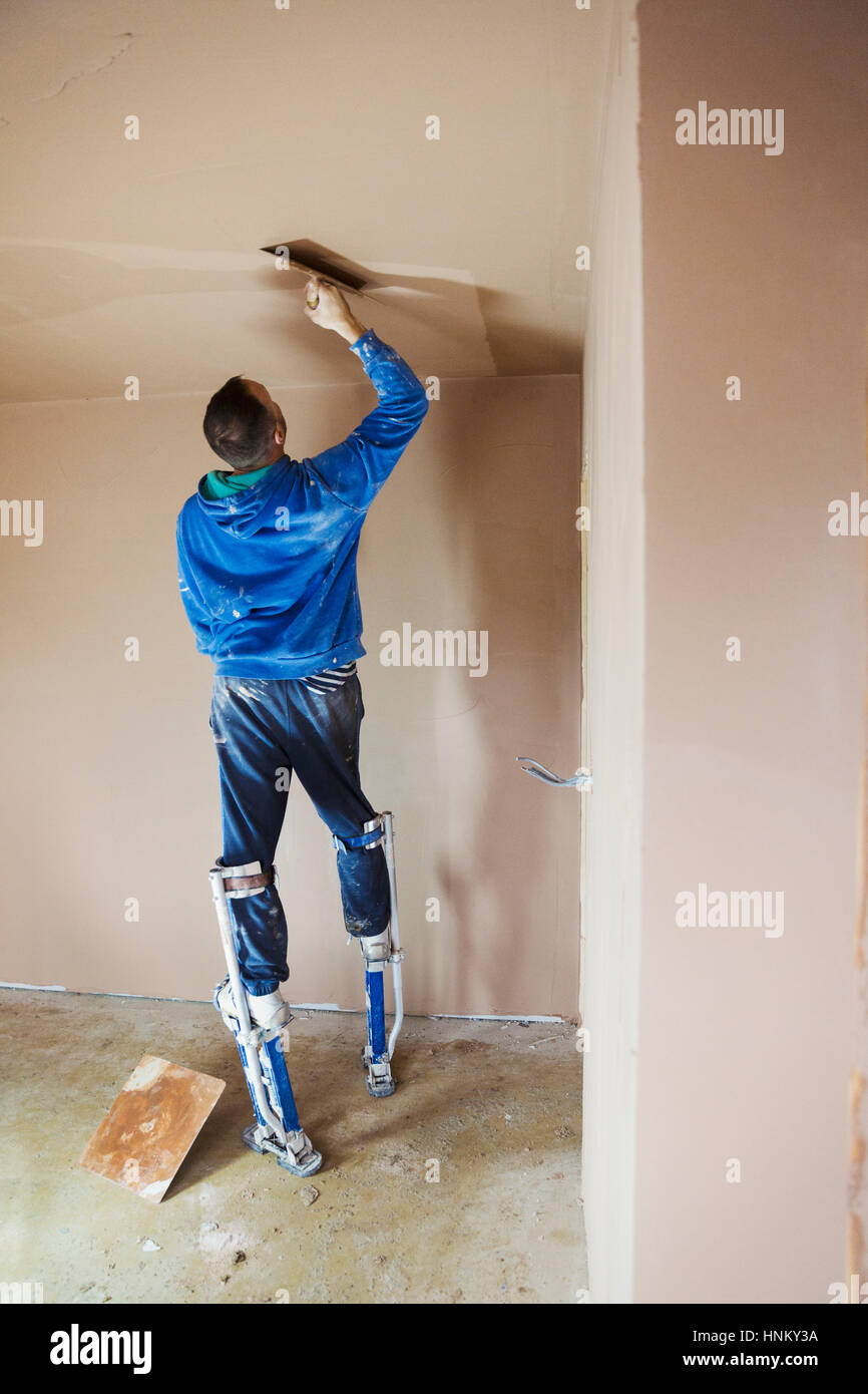 A plasterer wearing stilts smoothing fresh plaster high up on the walls ...