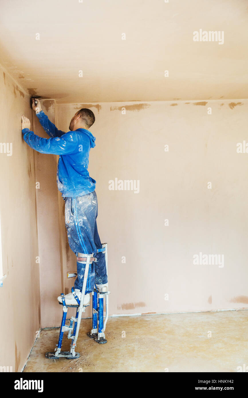 A plasterer wearing stilts smoothing fresh plaster high up on the walls ...