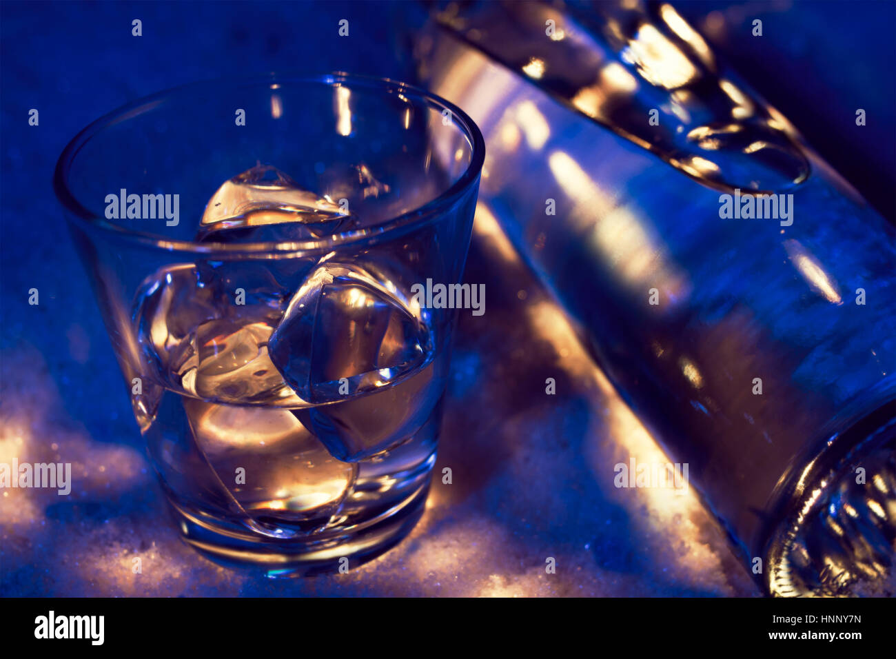 Bottle of vodka in the snow with glass filled with ice cubes, dark blue ...