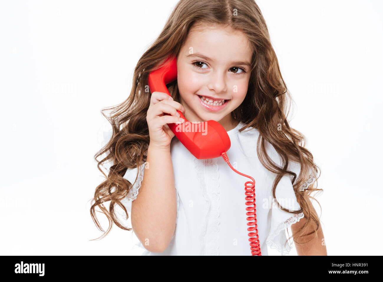 Cheerful cute little girl talking on red telephone over white ...