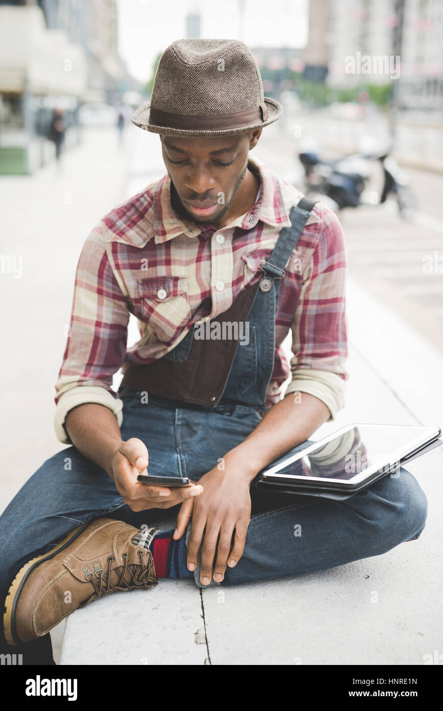 Young handsome afro black man sitting outdoor using smart phone and tablet - multitasking ...