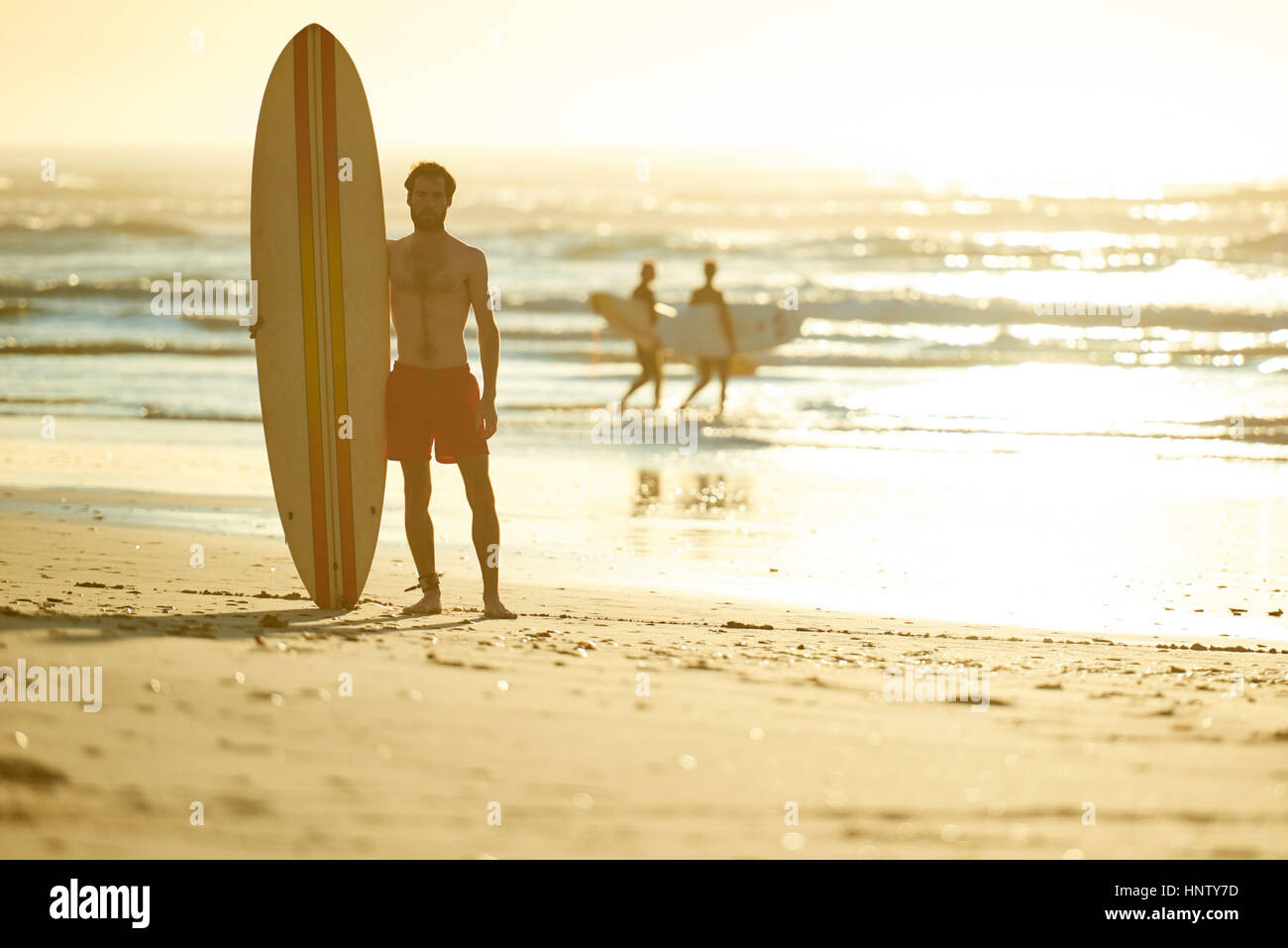 Surfer standing on beach with other surfers behind him Stock Photo - Alamy