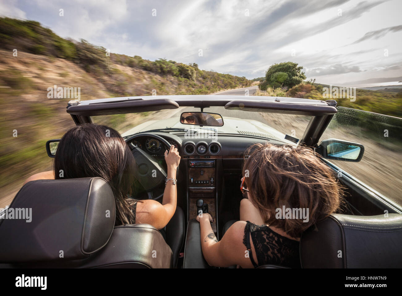 Two young women in convertible car, driving along scenic road, rear ...