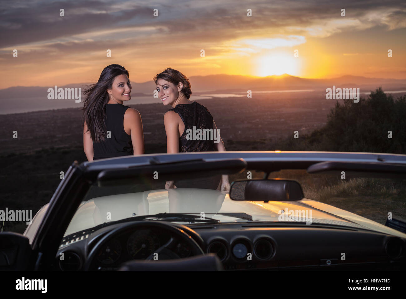 Two young sitting on bonnet of convertible car, looking over shoulder ...