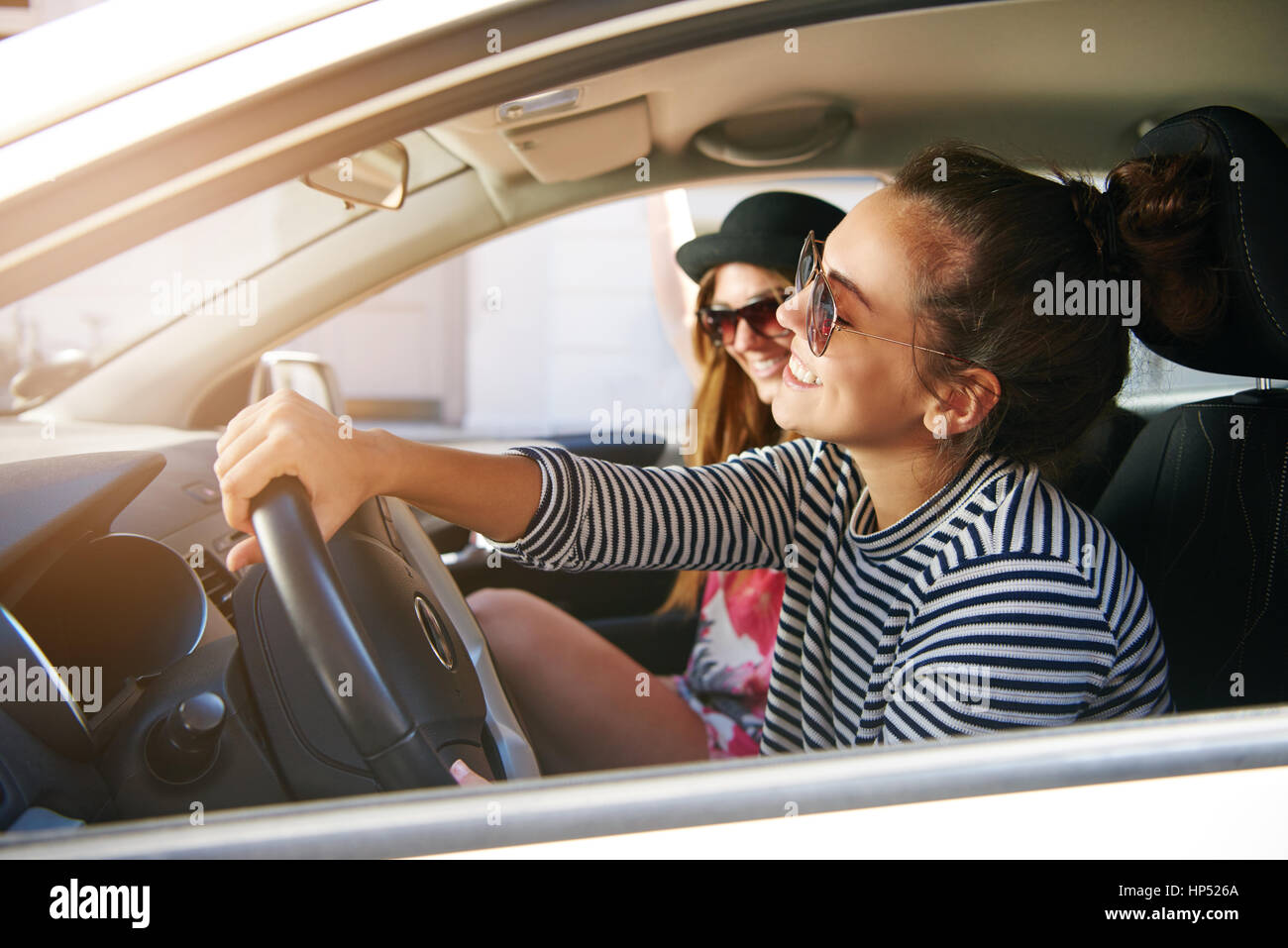 Pretty young woman driving a car with a friend smiling as they enjoy a ...