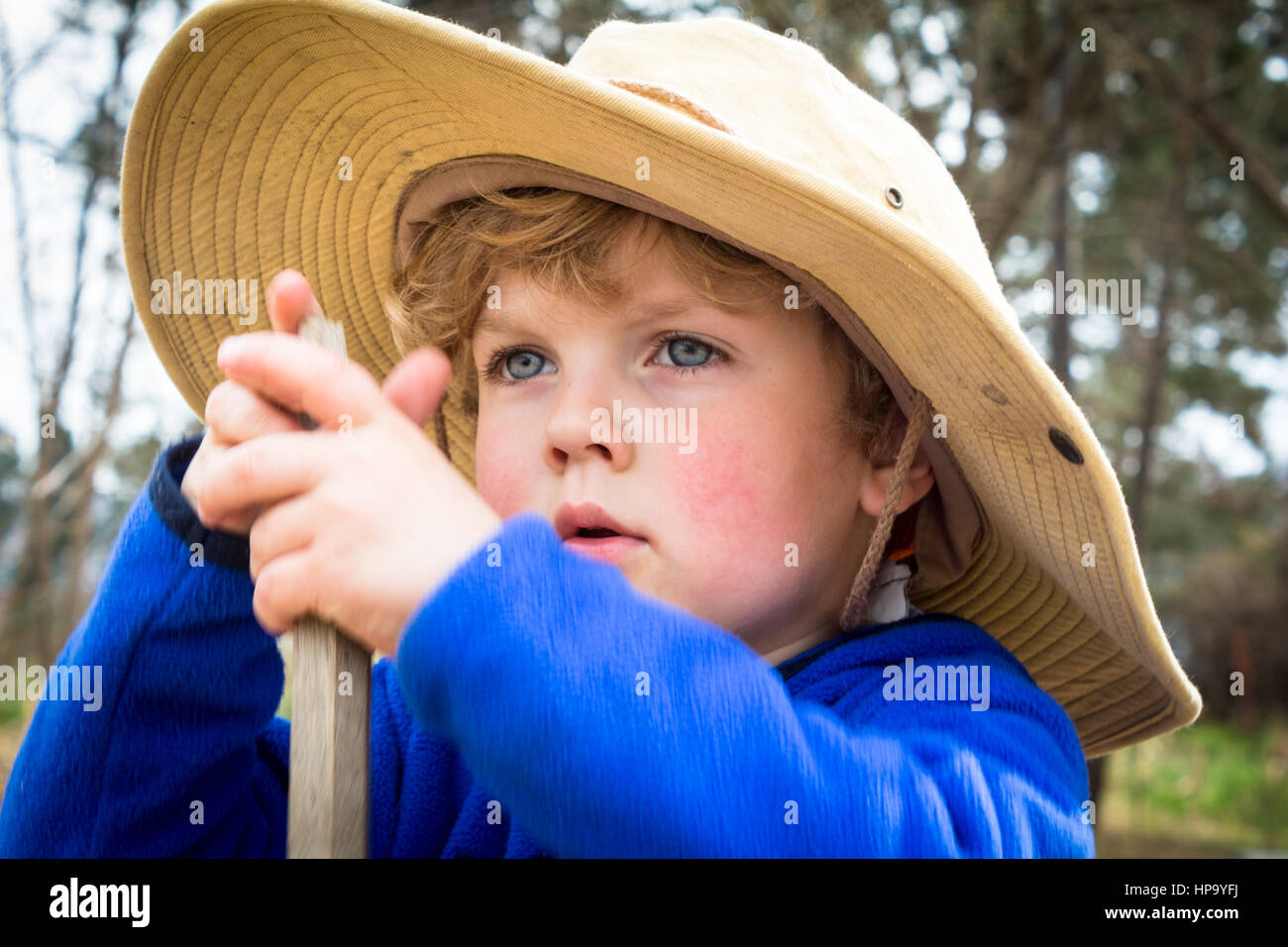 Pensive little farm boy with khaki hat playing with a stick outdoors ...