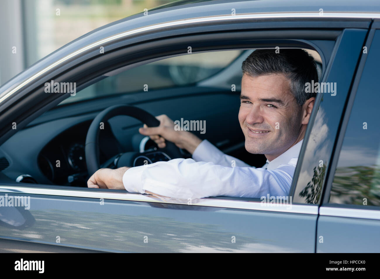 Confident businessman driving an expensive car, he is smiling at camera ...