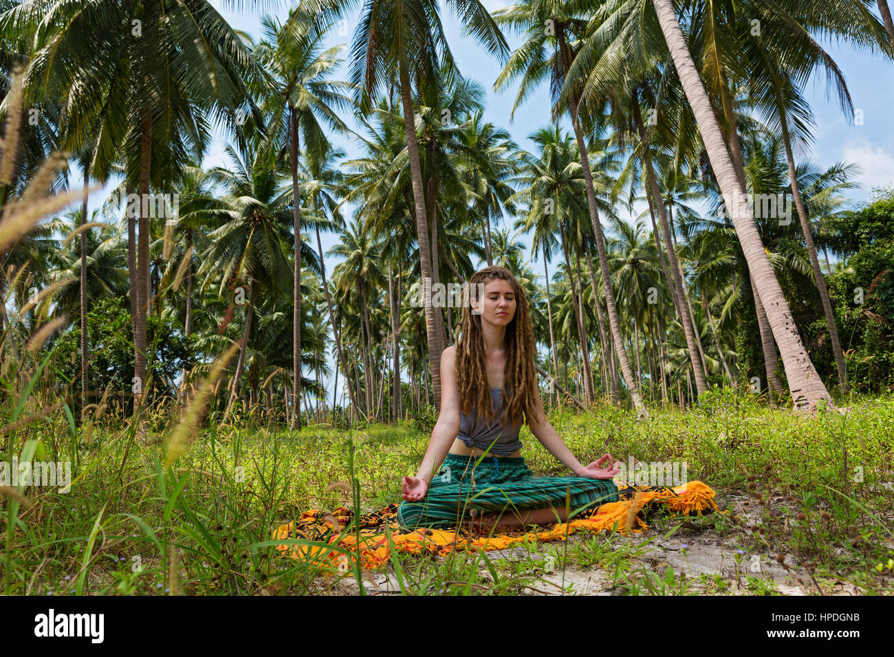 Young woman with dreadlocks doing yoga sitting amongst the palm trees ...