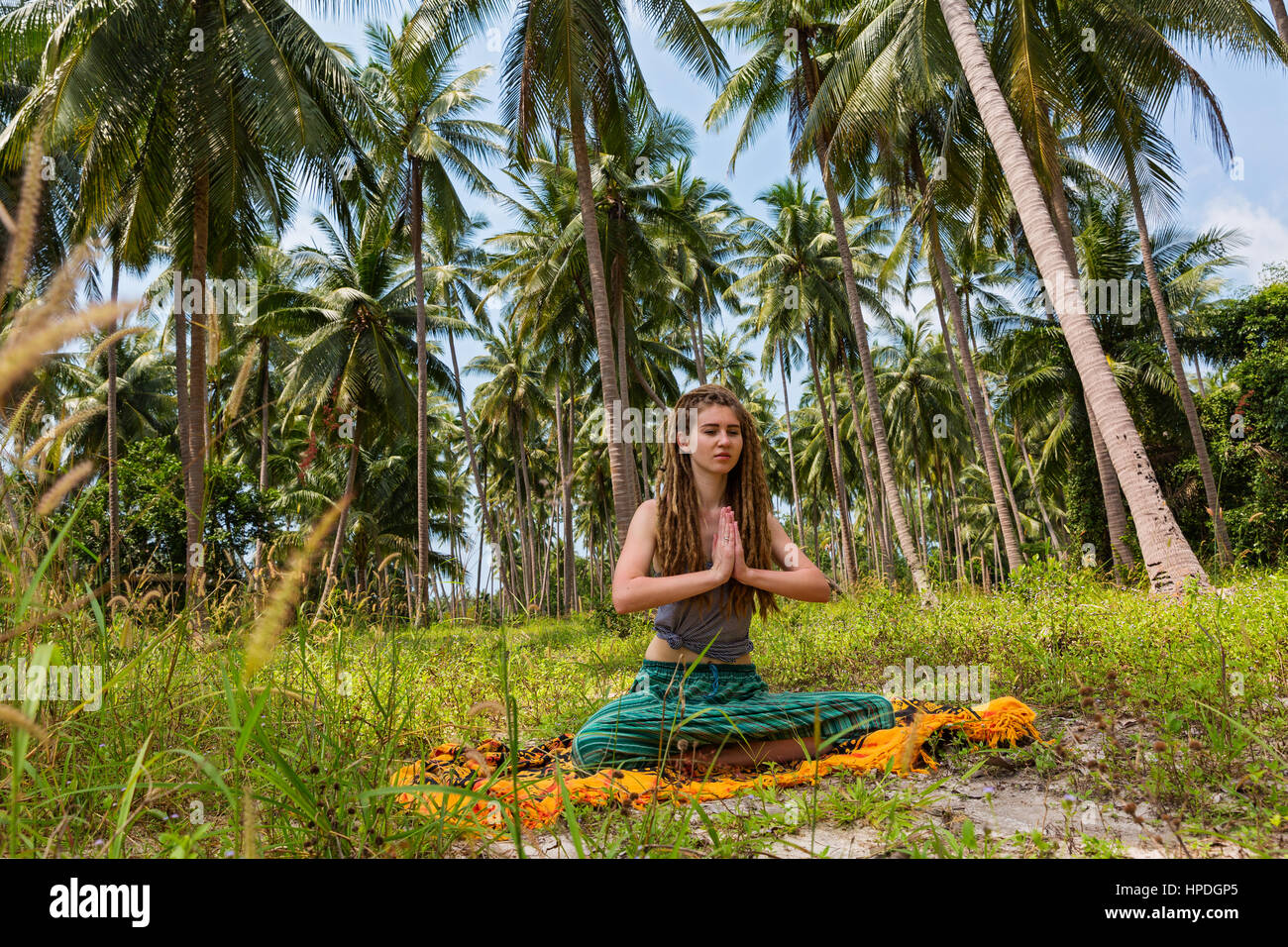 Young woman with dreadlocks doing yoga sitting amongst the palm trees ...