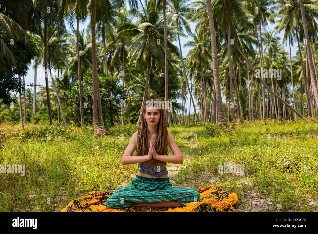 Young woman with dreadlocks doing yoga sitting amongst the palm trees ...