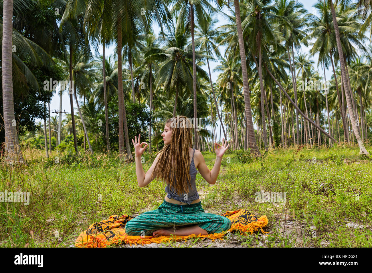 Young woman with dreadlocks doing yoga sitting amongst the palm trees ...
