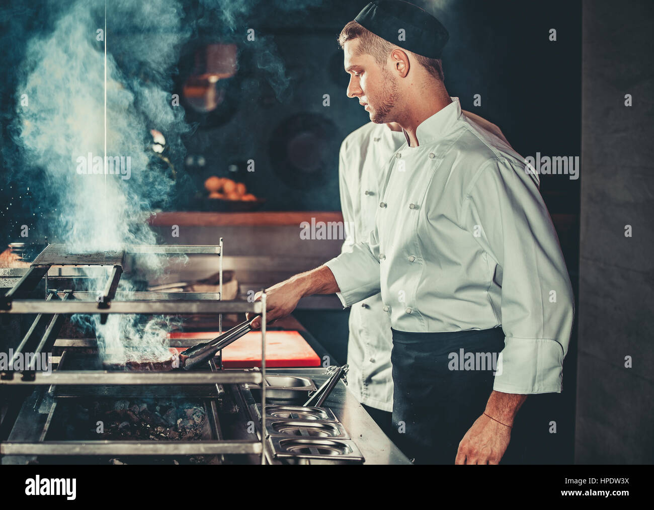 Young white chef in black apron and hat standing near the brazier whith ...