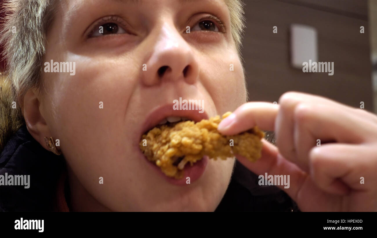 Woman expressively eats chicken in fast food restaurant. Face closeup ...