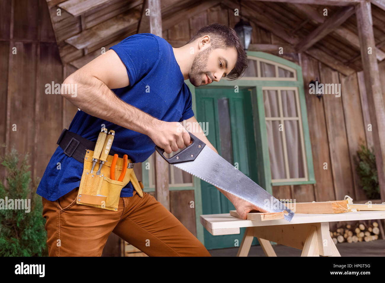 handsome bearded carpenter in tool belt sawing wooden plank on porch ...