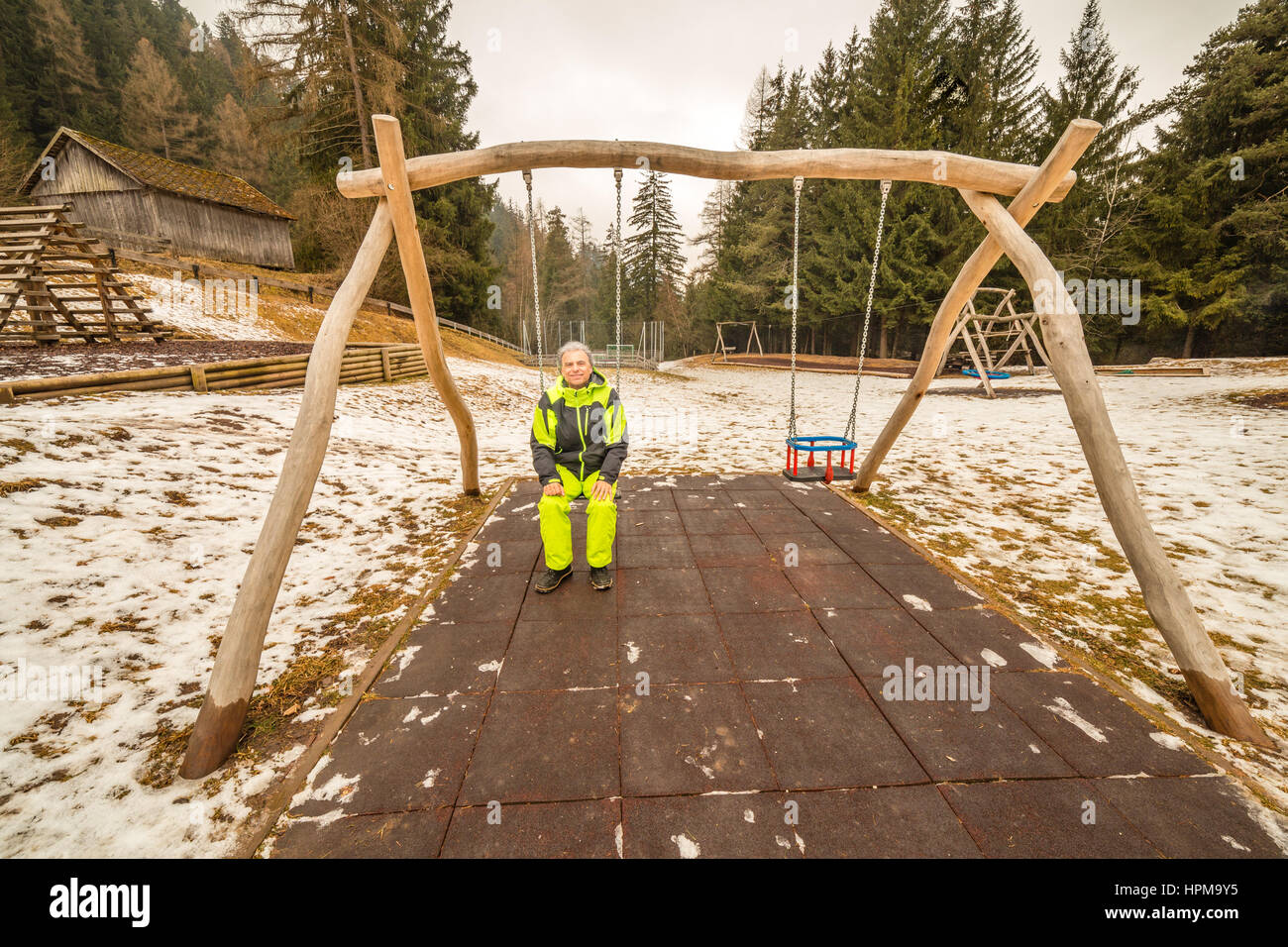 Happy mature man rocking on the swing in a snowy park in mountains ...