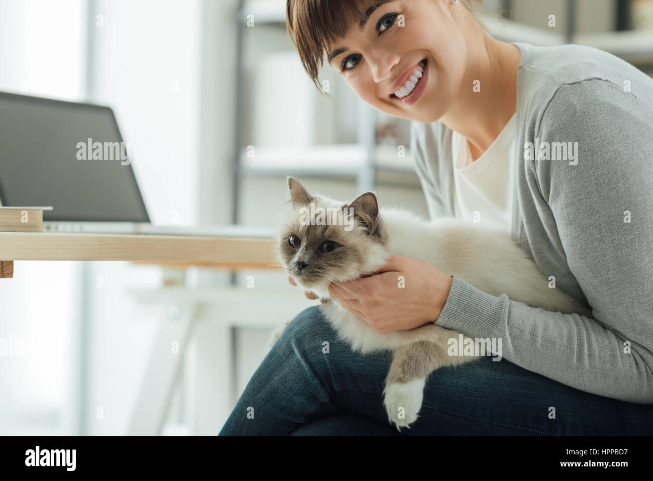 Happy smiling young woman at home cuddling her lovely cat on her legs ...