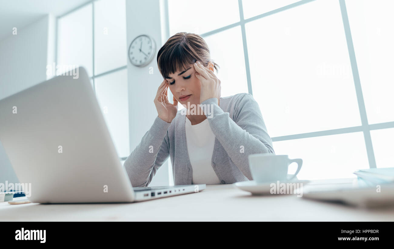 Exhausted young woman working at office desk touching her temples, she ...