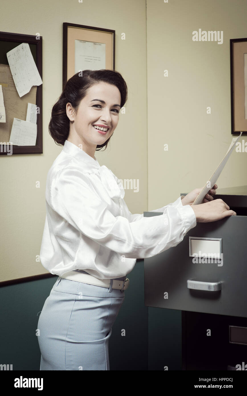 Smiling vintage secretary searching files in the filing cabinet drawers ...