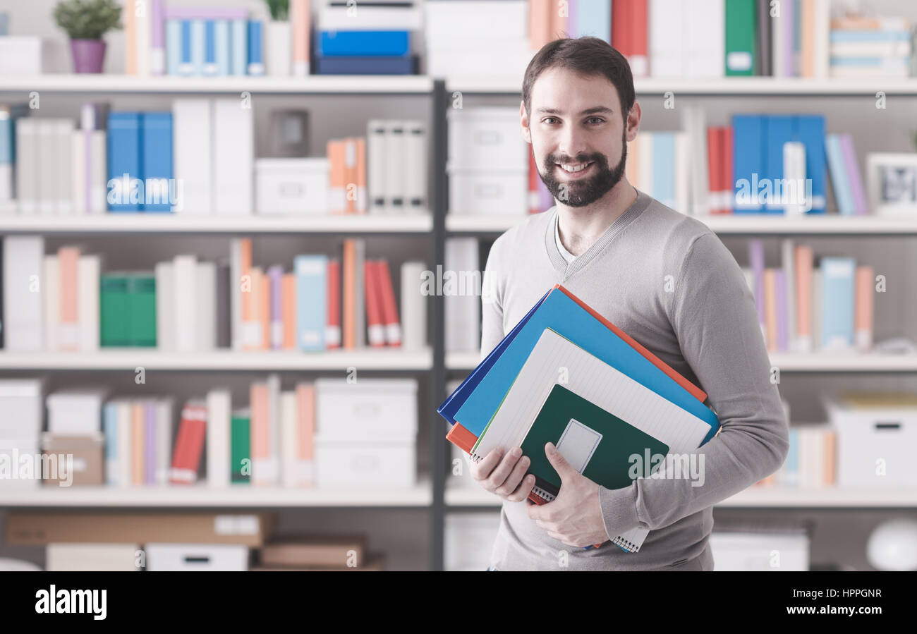 Man holding files and folders and smiling at camera, office shelves ...