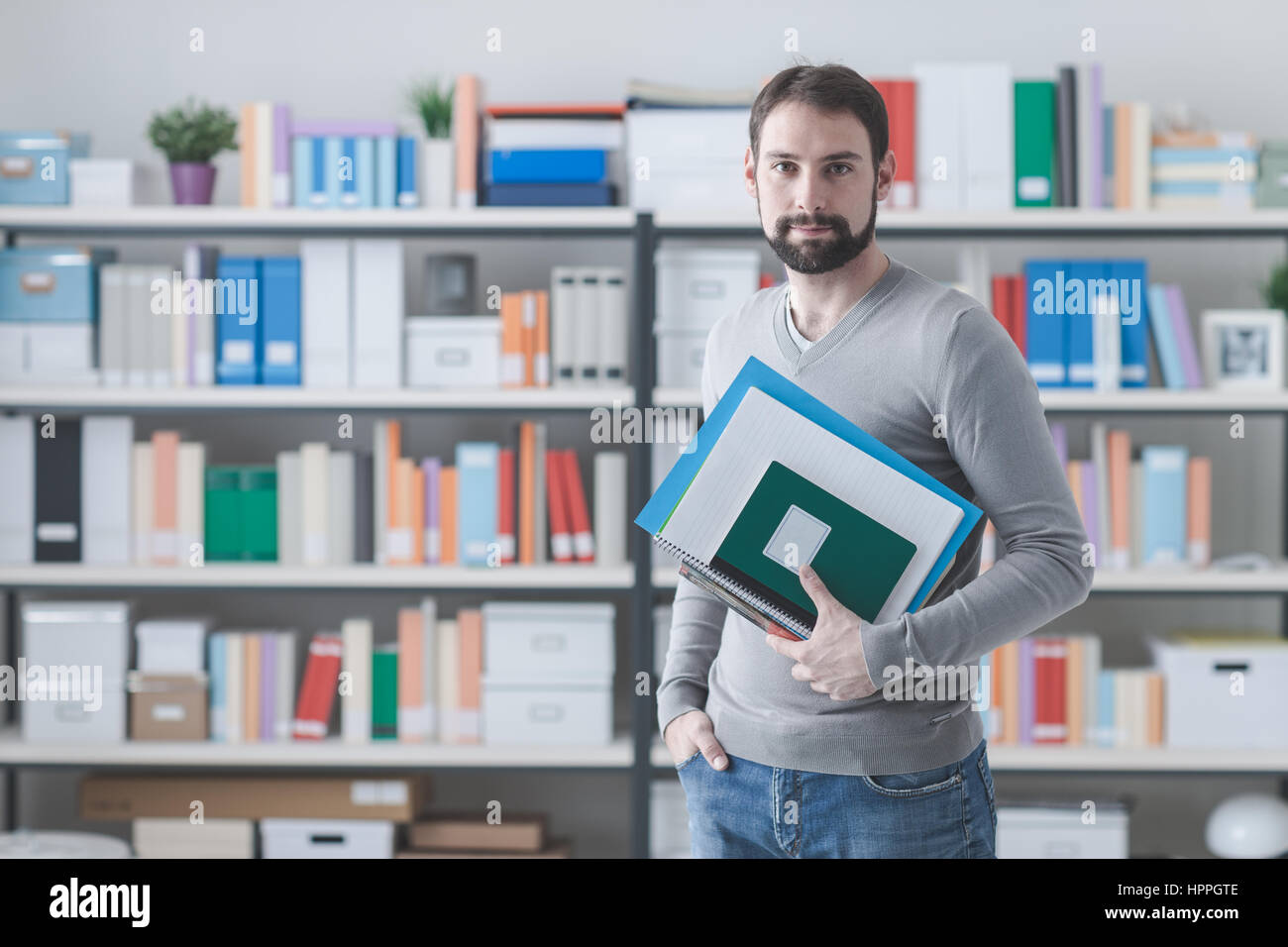 Man holding files and folders and smiling at camera, office shelves ...