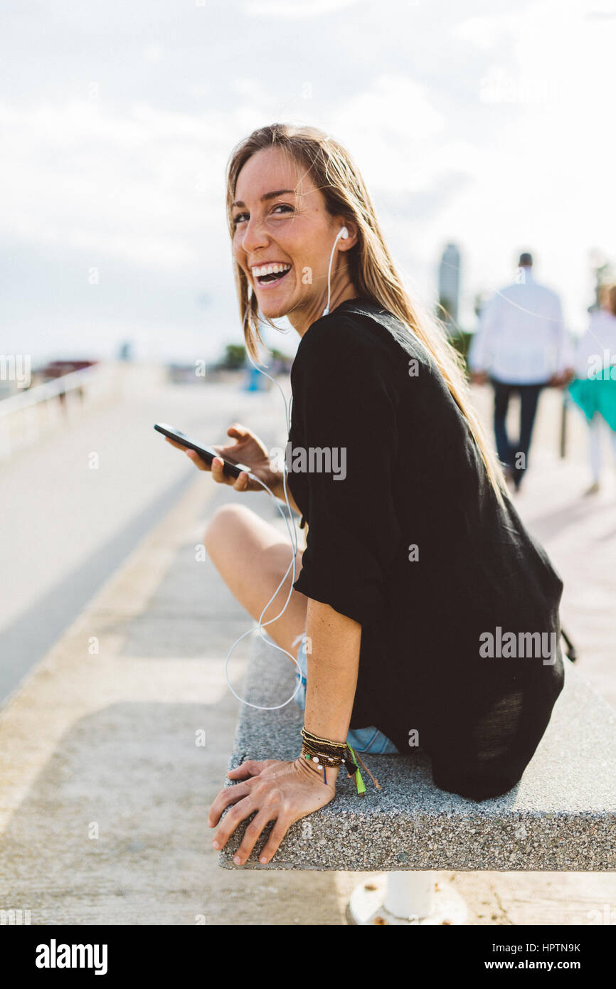 Happy young woman with cell phone and earbuds on waterfront promenade ...