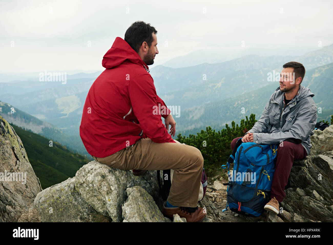 Two men in hiking gear sitting on a rock high in the mountains talking ...