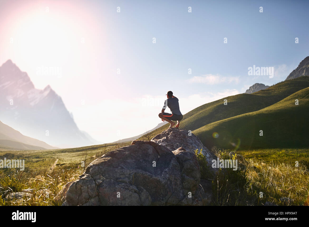 Young man crouching on rock, looking at sunny,r emote mountain view ...