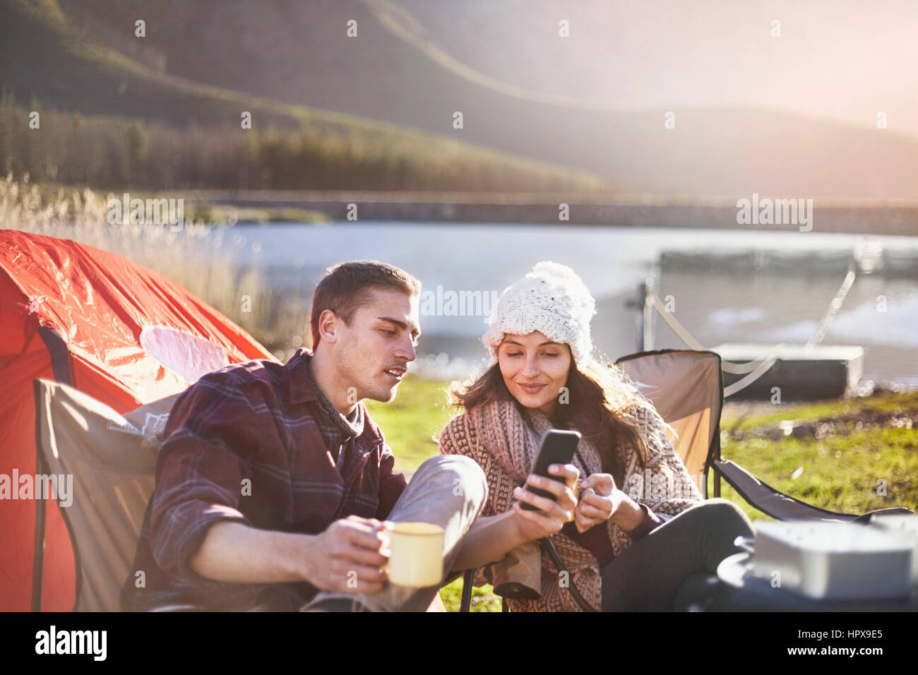 Young couple camping, drinking coffee and using cell phone at sunny lakeside campsite Stock ...