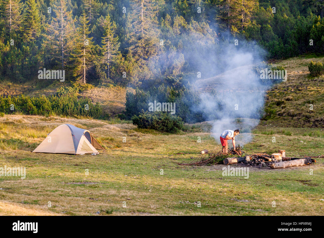 View with a tent, camping fire wit smoke and tourist hiker in summer ...