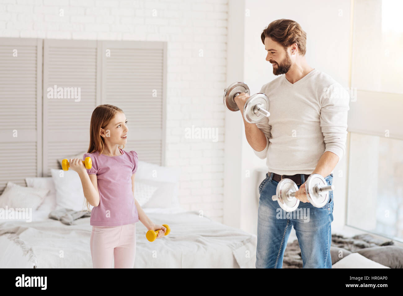 Keep fit. Sporty bearded man lifting bells standing near his daughter ...