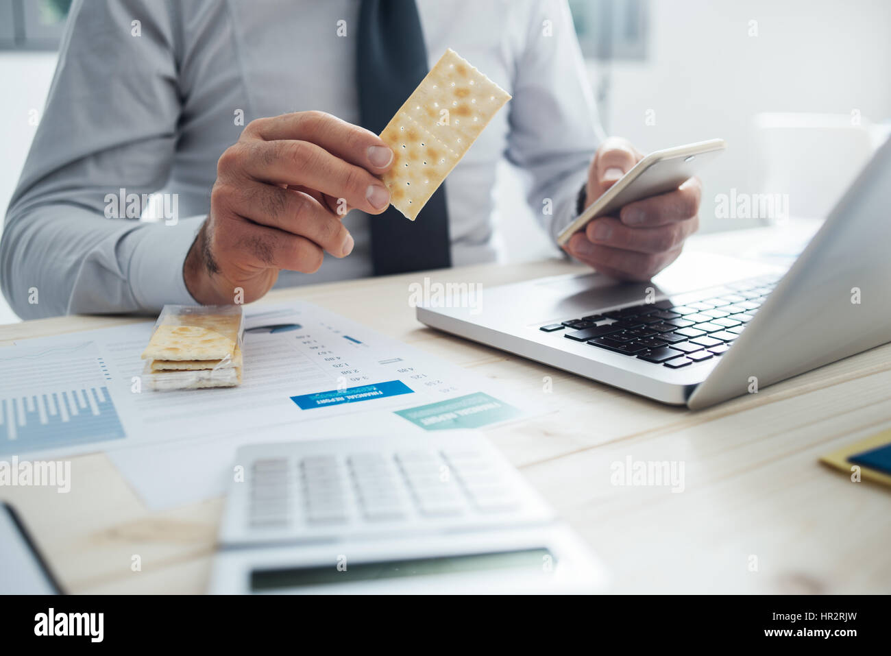 Businessman having a snack at desk holding a cracker and a smart phone ...