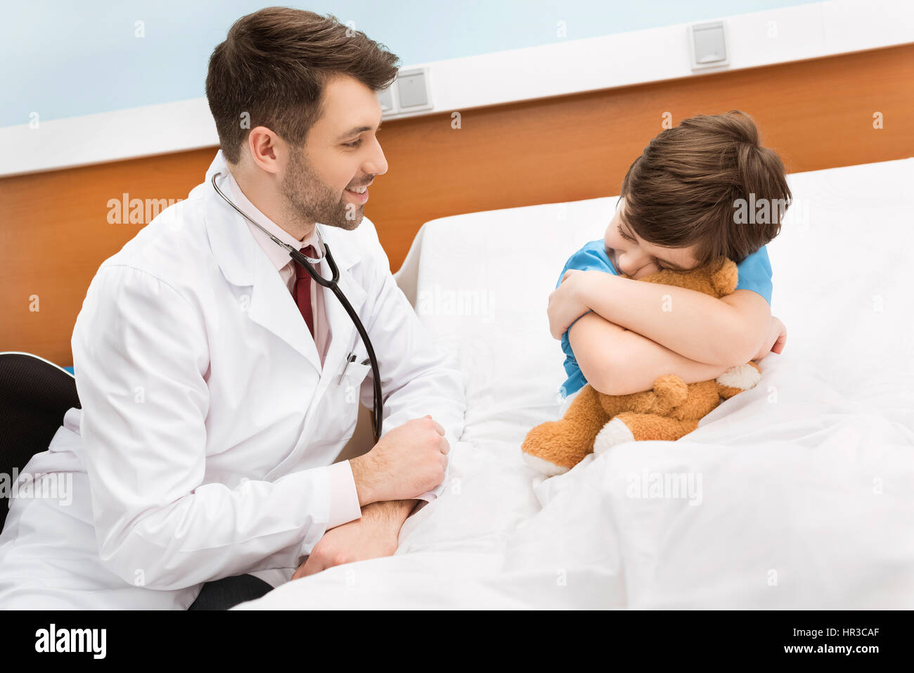 Smiling pediatrician looking at little boy hugging teddy bear in ...