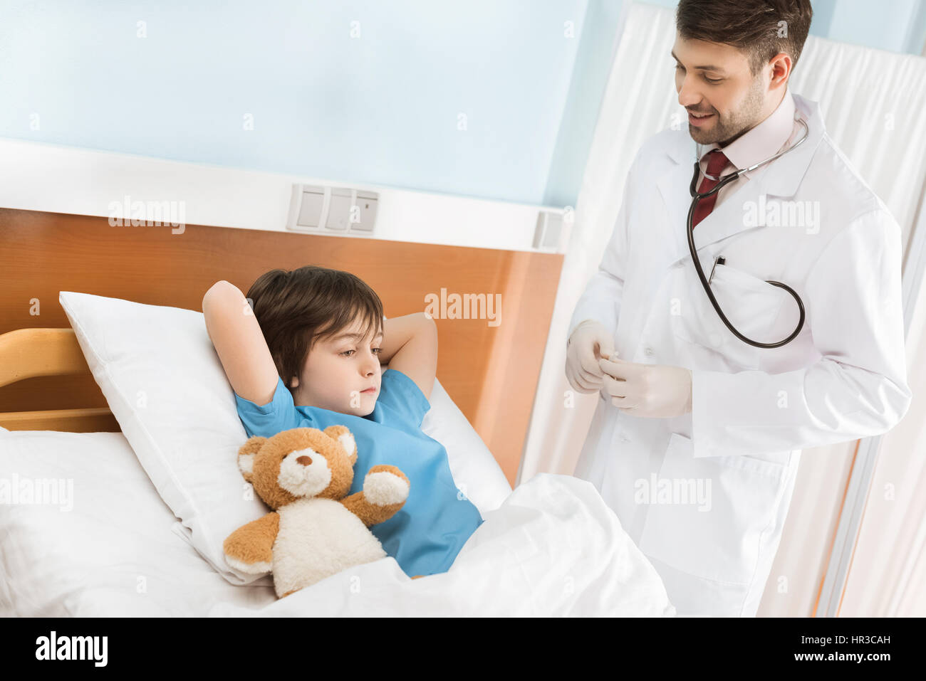 Smiling pediatrician looking at little boy lying with teddy bear in ...