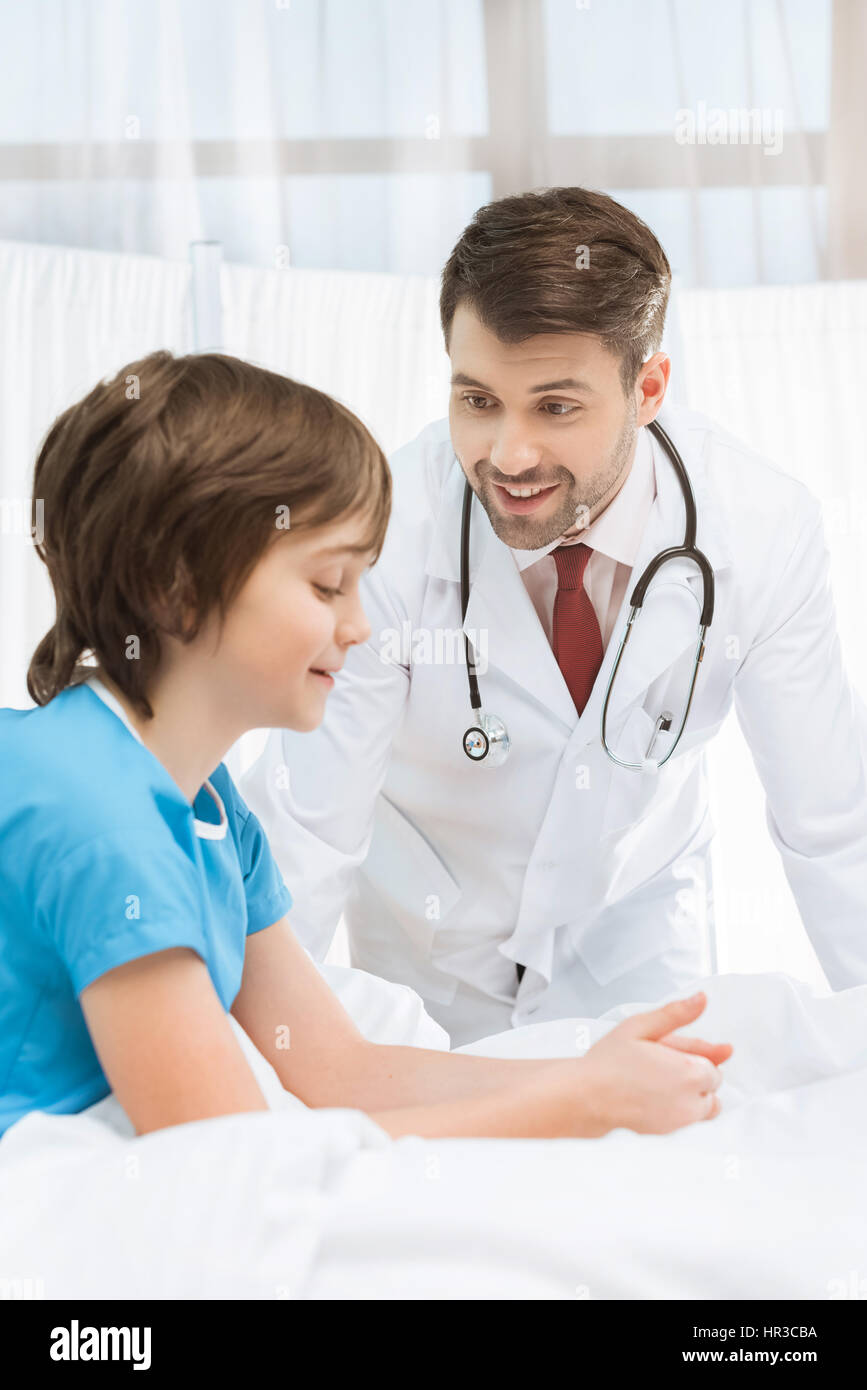 Smiling man doctor looking at little boy patient sitting on hospital ...