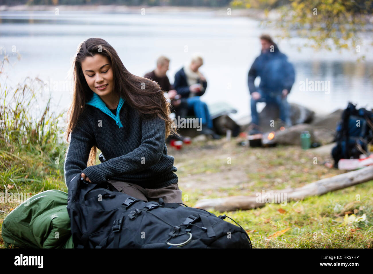 Young woman unpacking backpack with friends in background at campsite ...