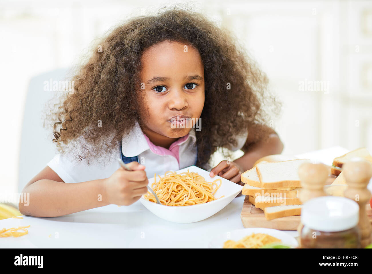 Curly little girl with full mouth looking at camera while chewing tasty ...