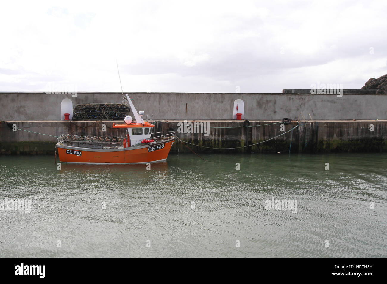Small orange coloured inshore fishing boat with rope moored alongside stone quay in the harbour ...