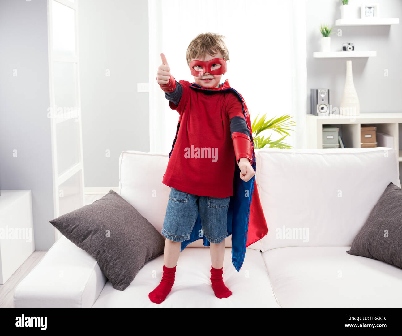 Smiling superhero boy standing on sofa with thumb up Stock Photo - Alamy