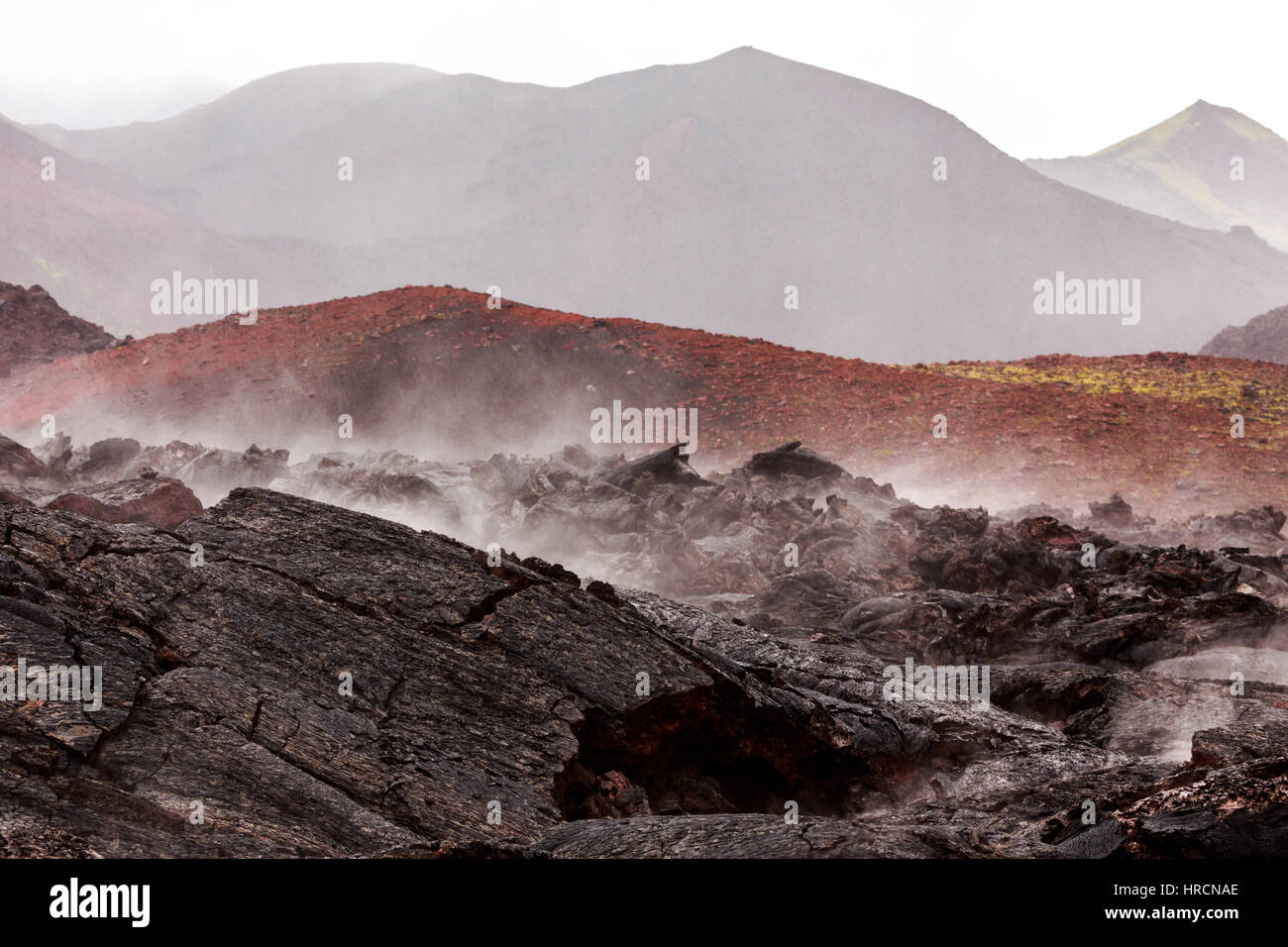 Barren lava fields steaming in light rain with volcanoes in background ...