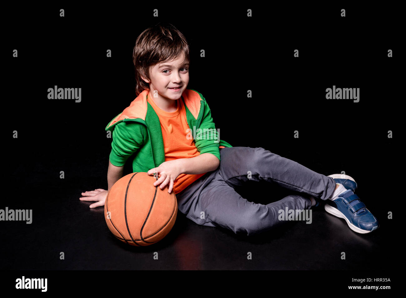 smiling boy sitting on floor with basketball ball and looking away on ...