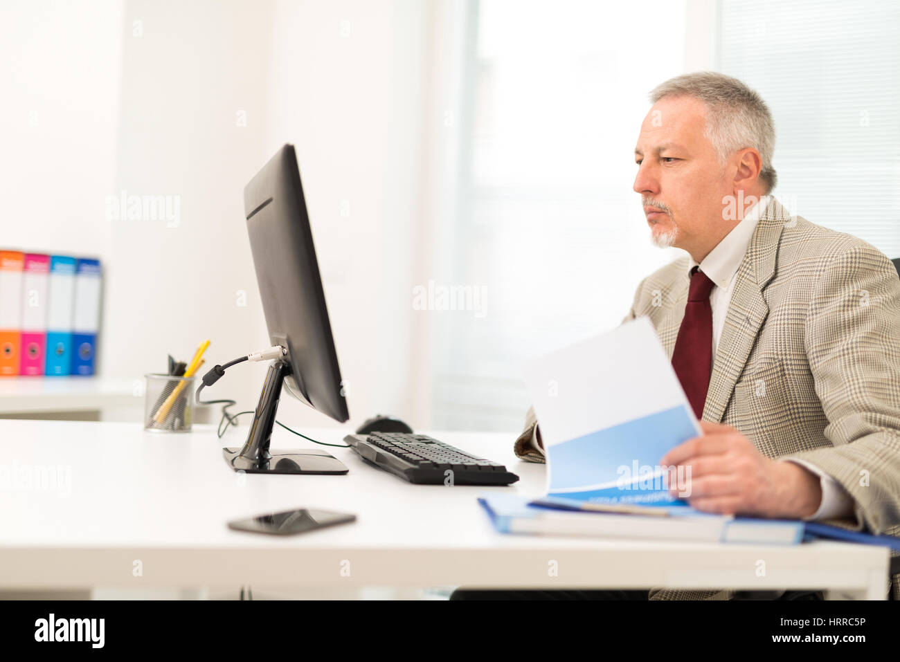 Senior businessman working on his computer while reading a document ...