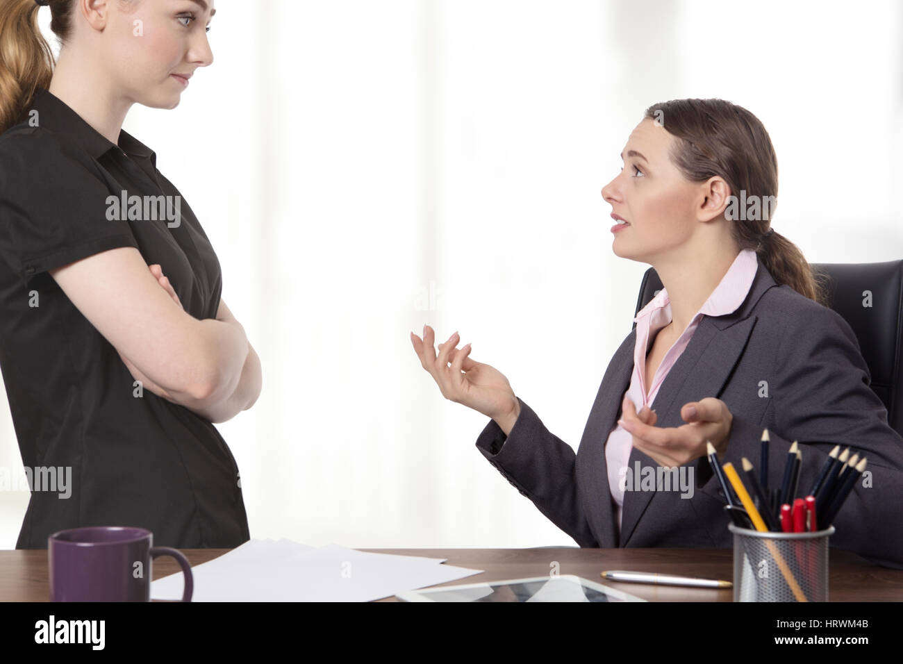 two young, female co-workers in an office. One sitting at a desk ...