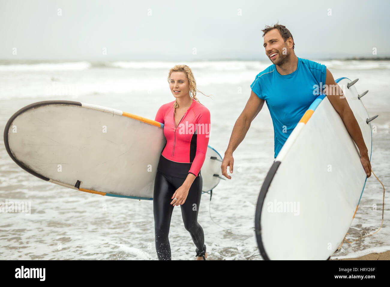 Joyful Couple Of Surfers Walks With Surfboards On The Beach On The