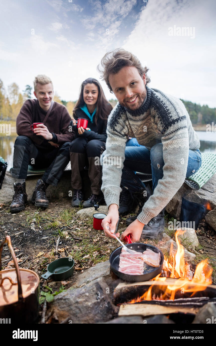 Portrait of young man cooking food on campfire with friends in ...